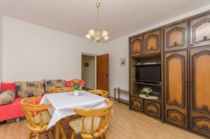 Uma área de refeições com uma mesa posta para quatro pessoas, cercada por cadeiras de madeira. Um lustre pendurado no teto, e uma televisão fixada em um armário de madeira.