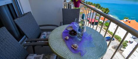 Uma varanda com móveis de vime, uma mesa com objetos decorativos e vista para uma cidade costeira e o mar.