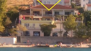 Um edifício costeiro com várias varandas, uma praia arenosa em primeiro plano e uma paisagem acidentada ao fundo.