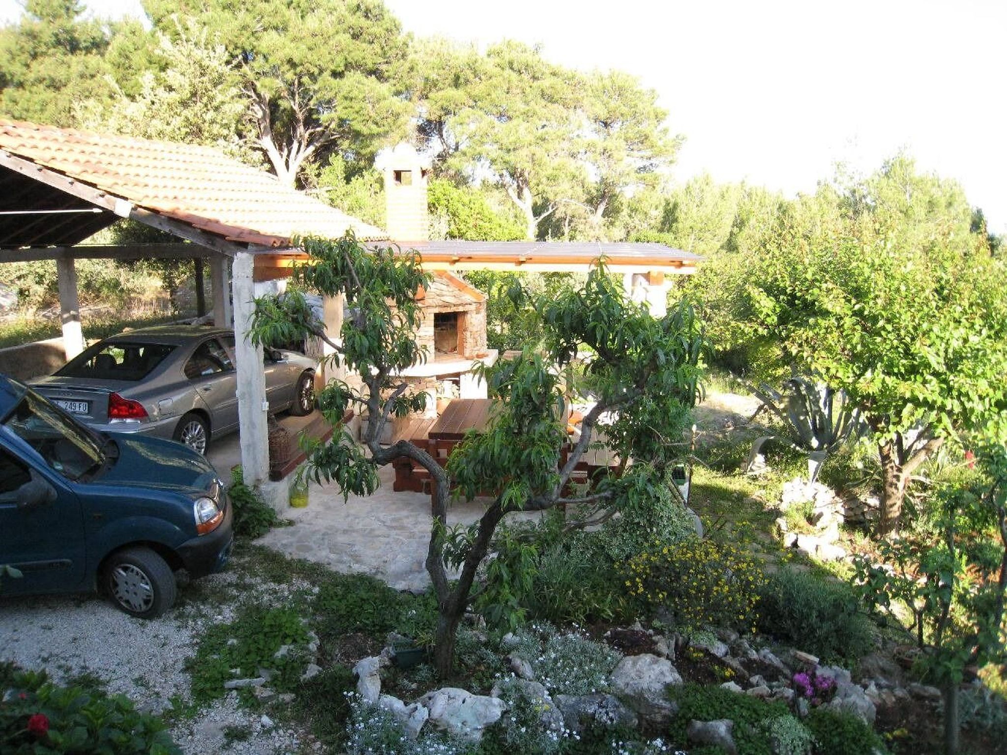 Uma área externa coberta com um carro estacionado sob uma cobertura, um edifício de pedra e um jardim com diversas plantas e flores.