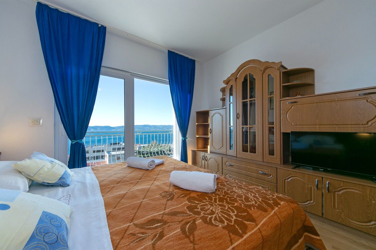 Quarto de hotel com uma cama grande, televisão, armários de madeira e varanda com vista para o mar.