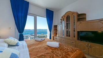 Quarto de hotel com uma cama grande, televisão, armários de madeira e varanda com vista para o mar.