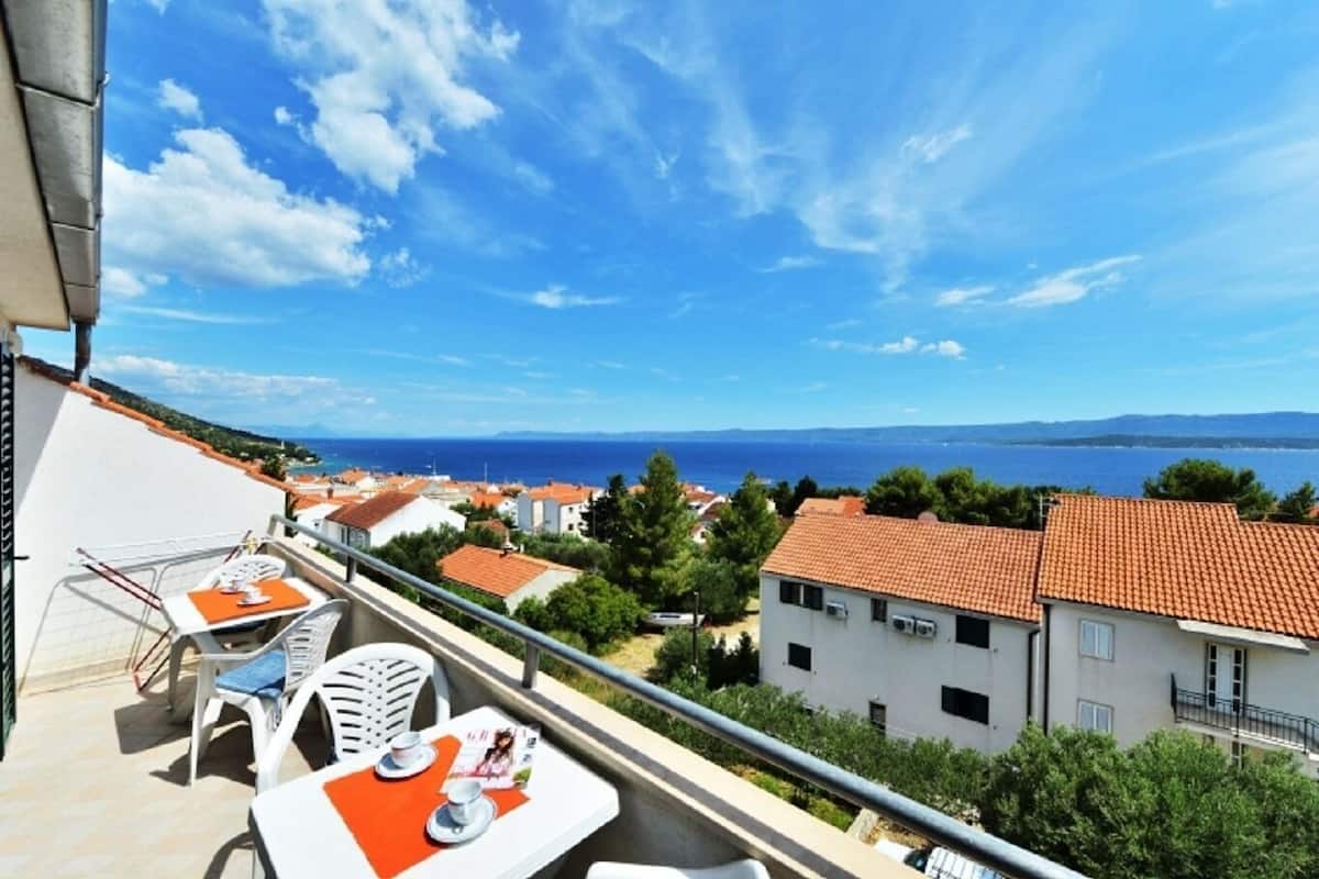 Varanda com mesa posta para dois, com vista para uma cidade costeira com edifícios de telhado vermelho e céu azul límpido.