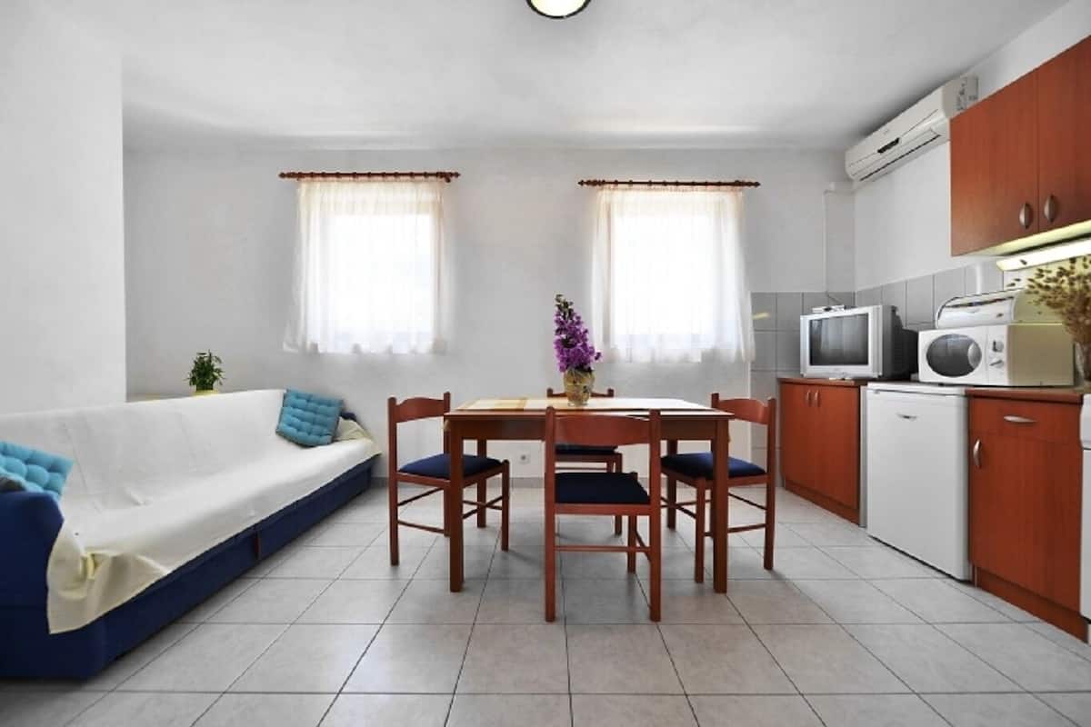Um espaço compacto para viver, com sofá, mesa de jantar, cadeiras, pequena cozinha e duas janelas com cortinas.