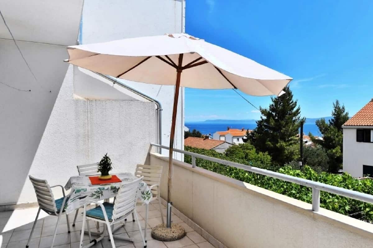 Uma varanda com mesa e cadeiras, um guarda-sol branco e vista para o mar e casas.