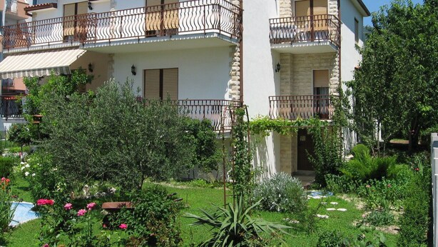 Uma casa de dois andares com varandas, um jardim com diversas plantas e flores, e um céu azul límpido.