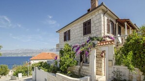 Uma casa de pedra de dois andares com telhado de telhas, um pequeno jardim com flores roxas e vista para o mar ao fundo.