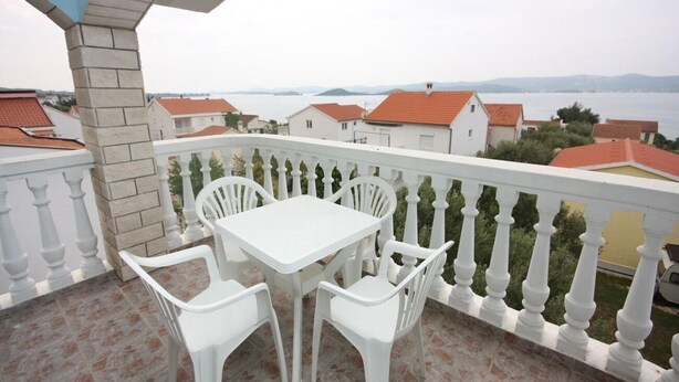 Uma varanda com mesa e cadeiras brancas, balaústres brancos e vista para casas e água ao fundo.