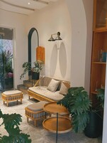 Uma sala de estar aconchegante com um sofá, dois pufes, uma mesa de centro e uma mesa lateral. Há uma planta grande, um abajur fixado na parede e um armário de madeira.