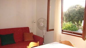 Quarto com sofá vermelho, cadeira de madeira, mesa com toalha branca, ventilador e janela com vista para vegetação.