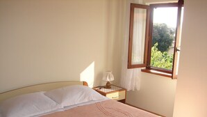 Cama de solteiro com colcha rosa e travesseiros brancos num quarto com janela, mesa de cabeceira com abajur e cortina branca.