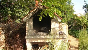 Forno de pedra ao ar livre com telhado inclinado, rodeado por vegetação e um caminho de terra.