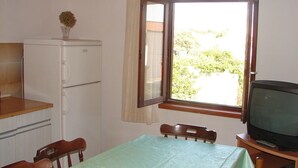 Um quarto pequeno com teto de madeira, geladeira branca, mesa com toalha verde, cadeira de madeira, televisão sobre um suporte de madeira e uma janela com cortina.