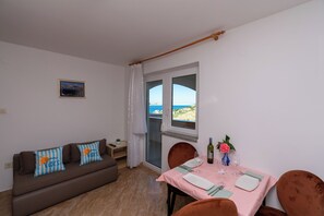 Quarto com sofá, mesa de jantar posta com pratos, copos e uma garrafa, e vista para o mar pela janela.