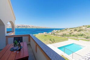 Uma varanda com mesa, piscina e vista para o mar e colinas.