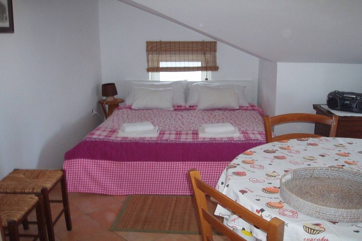 Quarto com uma cama, duas cadeiras, uma mesinha, uma janela com persianas e um rádio em cima de uma mesa.