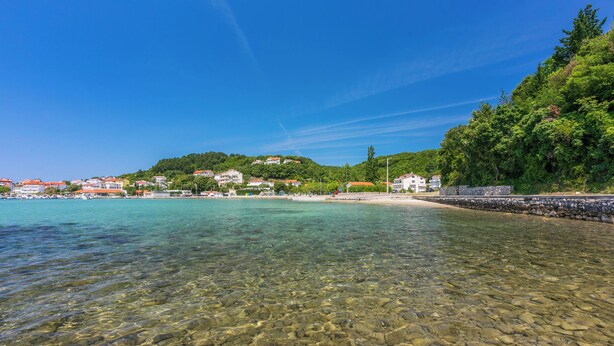 Uma área costeira com águas cristalinas, uma praia de areia e edifícios ao fundo.