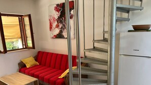 Um espaço de estar compacto com um sofá vermelho, uma pequena mesa de madeira, uma escada espiral de metal e um refrigerador branco.
