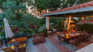 Um pátio externo com mesa e cadeiras de madeira, cercado por plantas em vasos e uma pérgola com telhado de azulejos.
