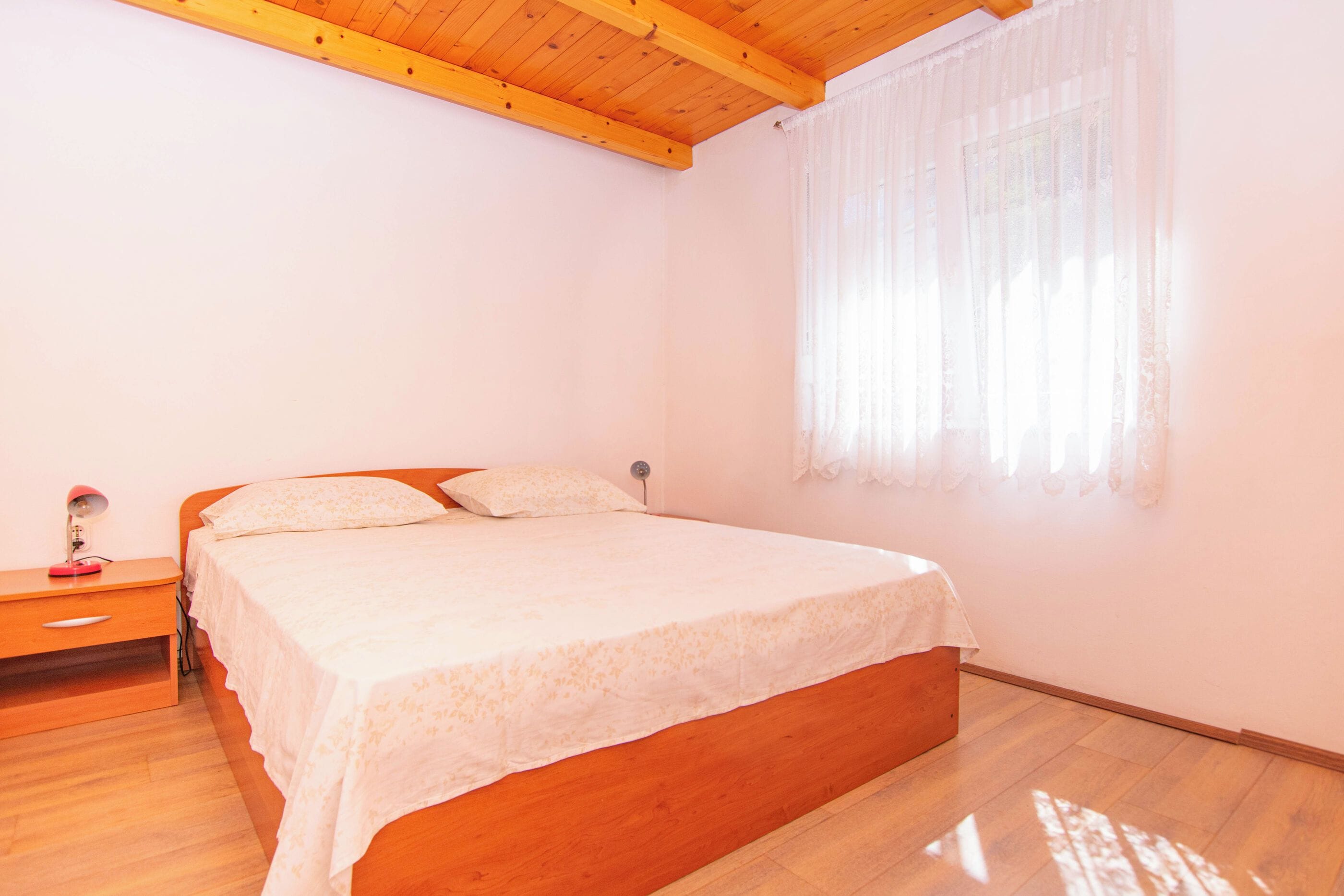 Quarto com cama de madeira, mesa de cabeceira, luminária e janela com cortinas leves.
