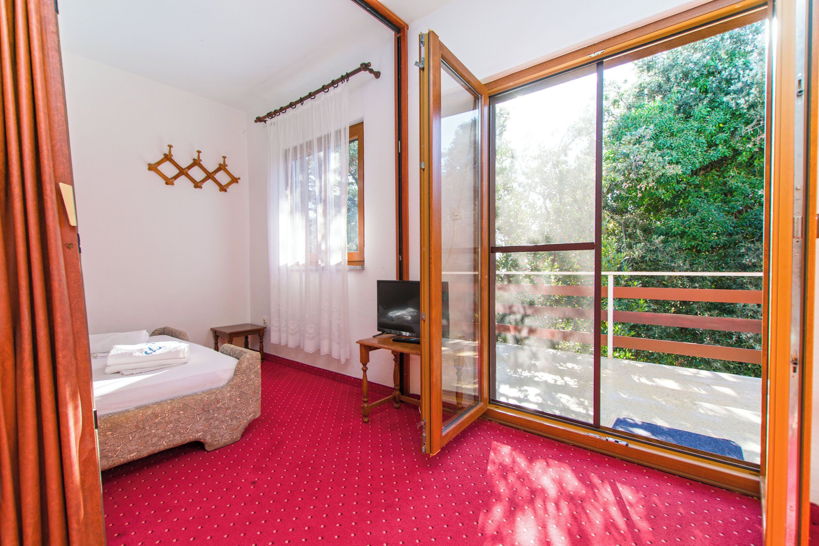 Quarto com cama, televisão, mesa pequena, janela com vista para o verde e tapete vermelho.