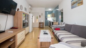 Uma sala de estar moderna com televisão de tela plana, mesa de centro de madeira, sofá e área de cozinha ao fundo.