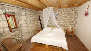 Um quarto com parede de pedra, cama com roupa de cama branca, cadeira de madeira, mesa de cabeceira com abajur e janela com cortina vermelha.