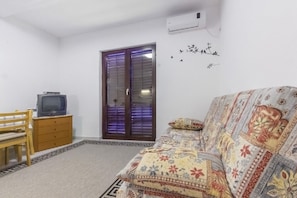 Quarto com sofá, uma cadeira de madeira, uma pequena TV em um armário de madeira, tapete estampado e uma porta de vidro deslizante com iluminação roxa no interior.