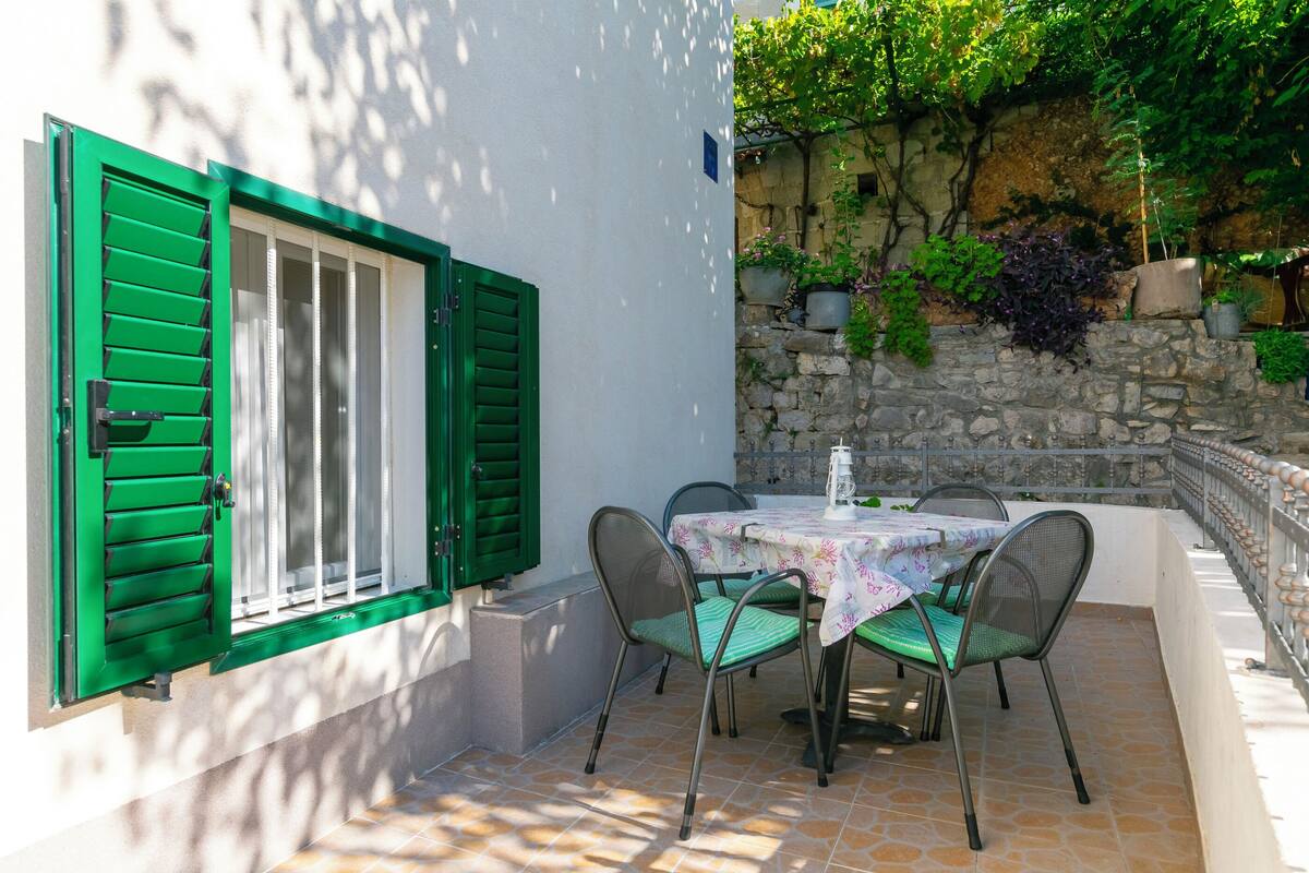 Um pátio com mesa e cadeiras, um muro de pedra com plantas e uma janela com persianas verdes.