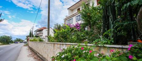 Rua residencial com casas, um muro de pedra e plantas floridas.