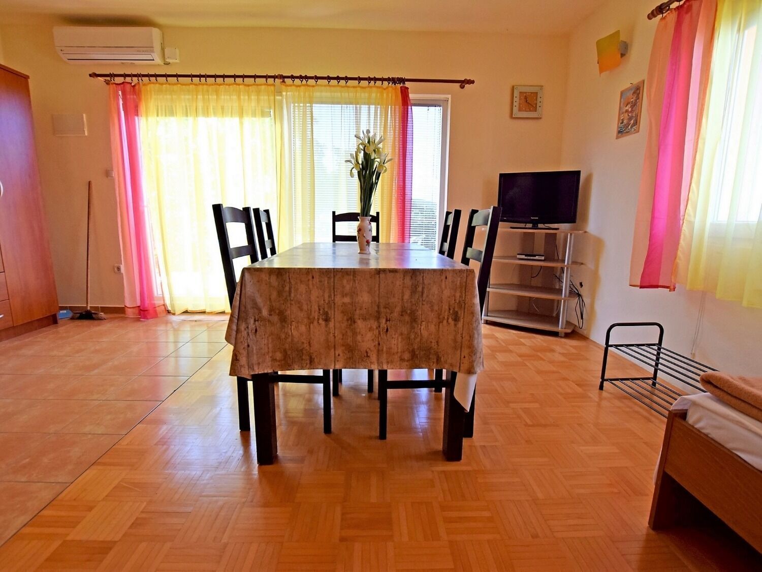 Uma sala de jantar com mesa, cadeiras, um vaso com flores, uma TV e uma cama.