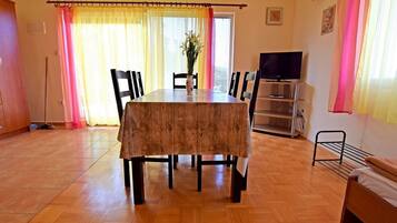 Uma sala de jantar com mesa, cadeiras, um vaso com flores, uma TV e uma cama.