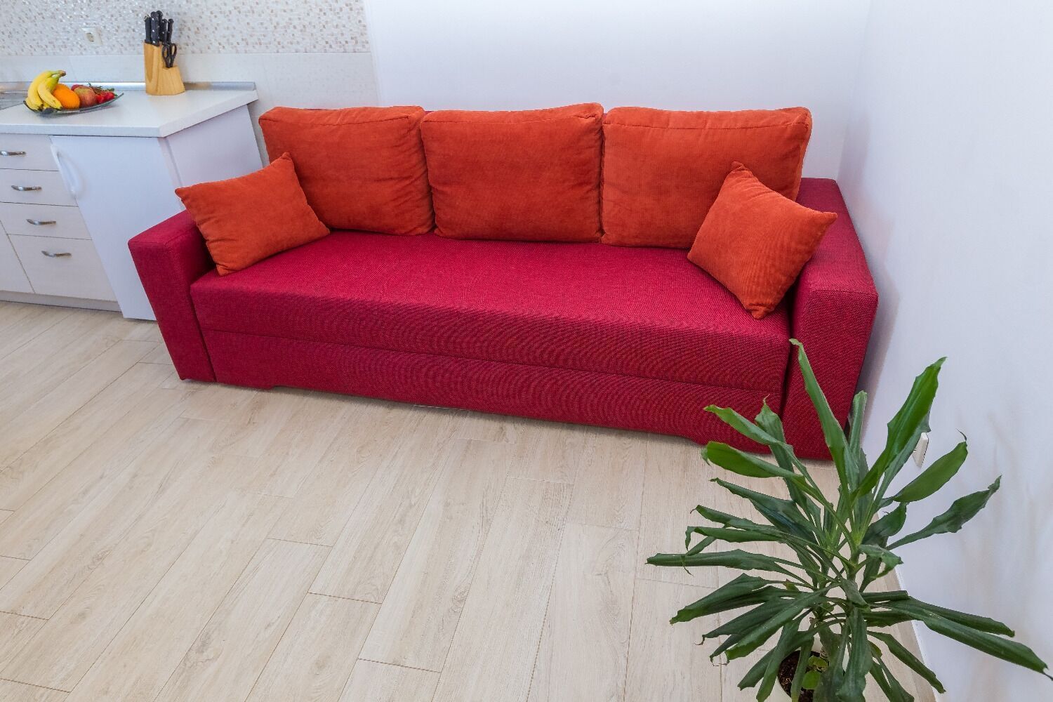 Um sofá vermelho com almofadas combinando em um quarto com piso de madeira clara e um balcão branco com frutas e utensílios.
