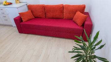 Um sofá vermelho com almofadas combinando em um quarto com piso de madeira clara e um balcão branco com frutas e utensílios.