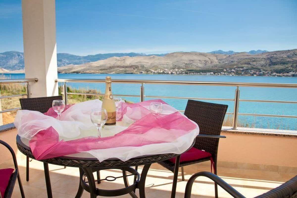Mesa posta para dois, com uma garrafa e copos, com vista para um lago e montanhas ao fundo.
