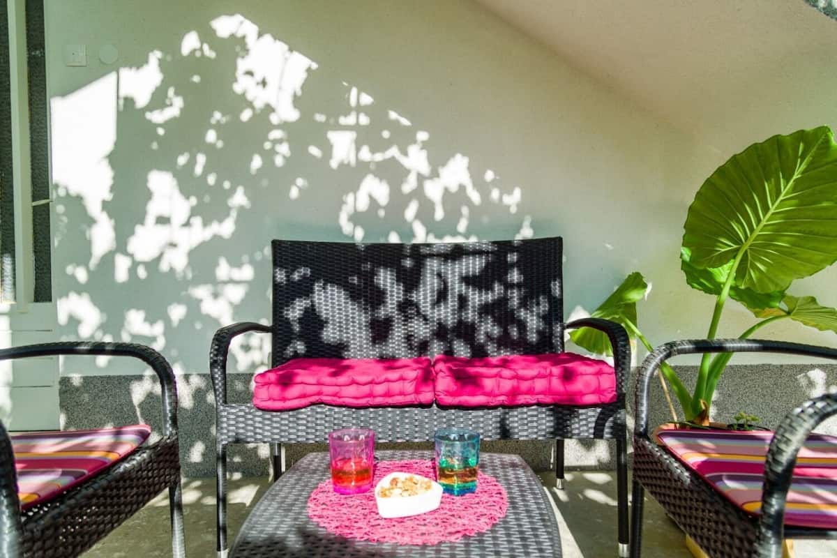 Um pátio com uma cadeira de rede preta com almofada rosa, dois copos de vidro e um pequeno prato de comida sobre um descanso de tecido.