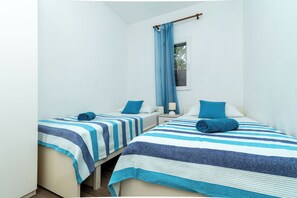 Quarto com duas camas, cada uma com colcha azul e branca listrada, uma janela com cortina e uma mesa de cabeceira com abajur.