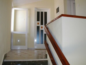 Um corredor com piso ladrilhado, corrimão de madeira e porta com painel de vidro.