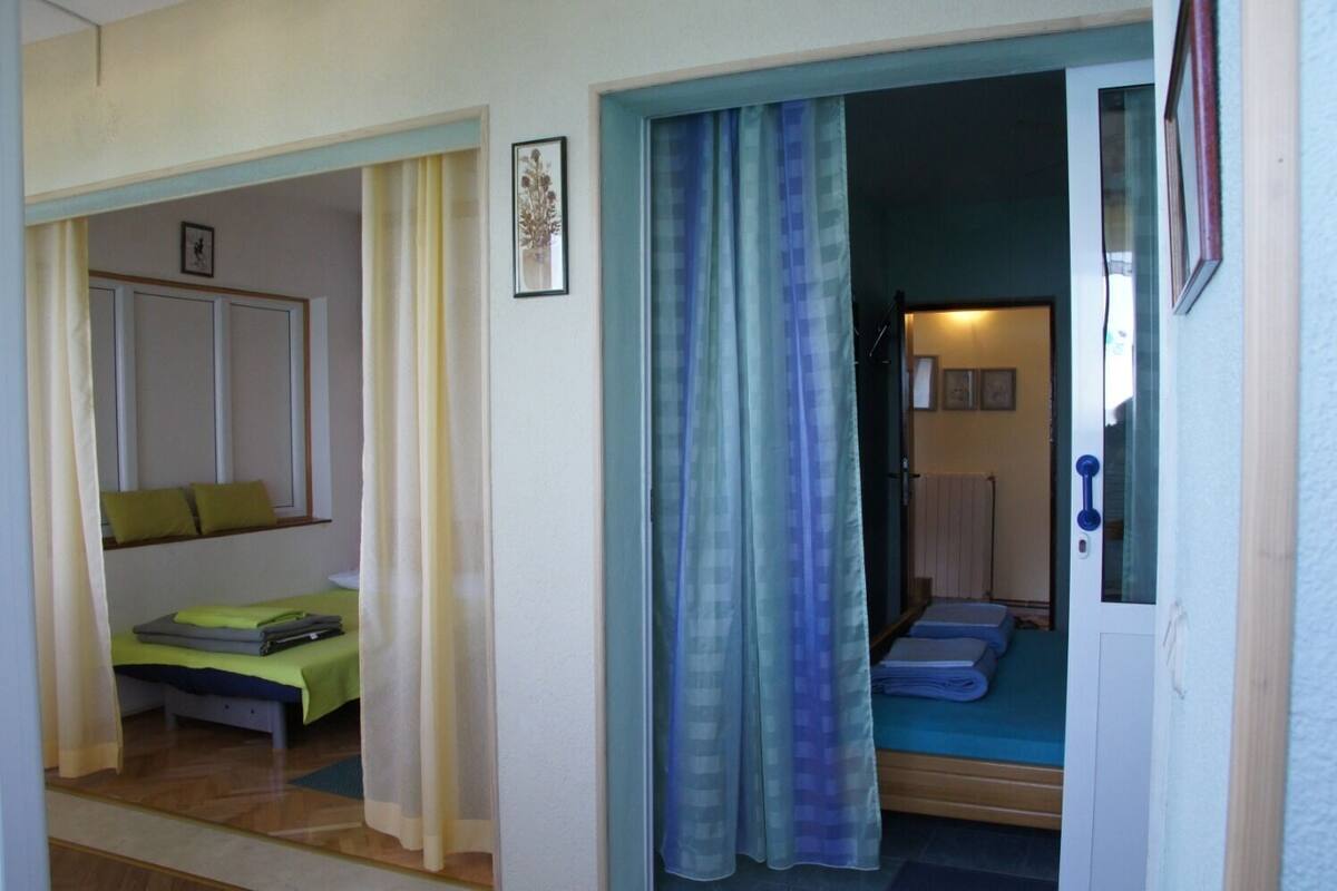 Quarto de hotel com uma cama, escrivaninha e uma cadeira. Há uma janela com cortinas e uma porta que dá para outro quarto.