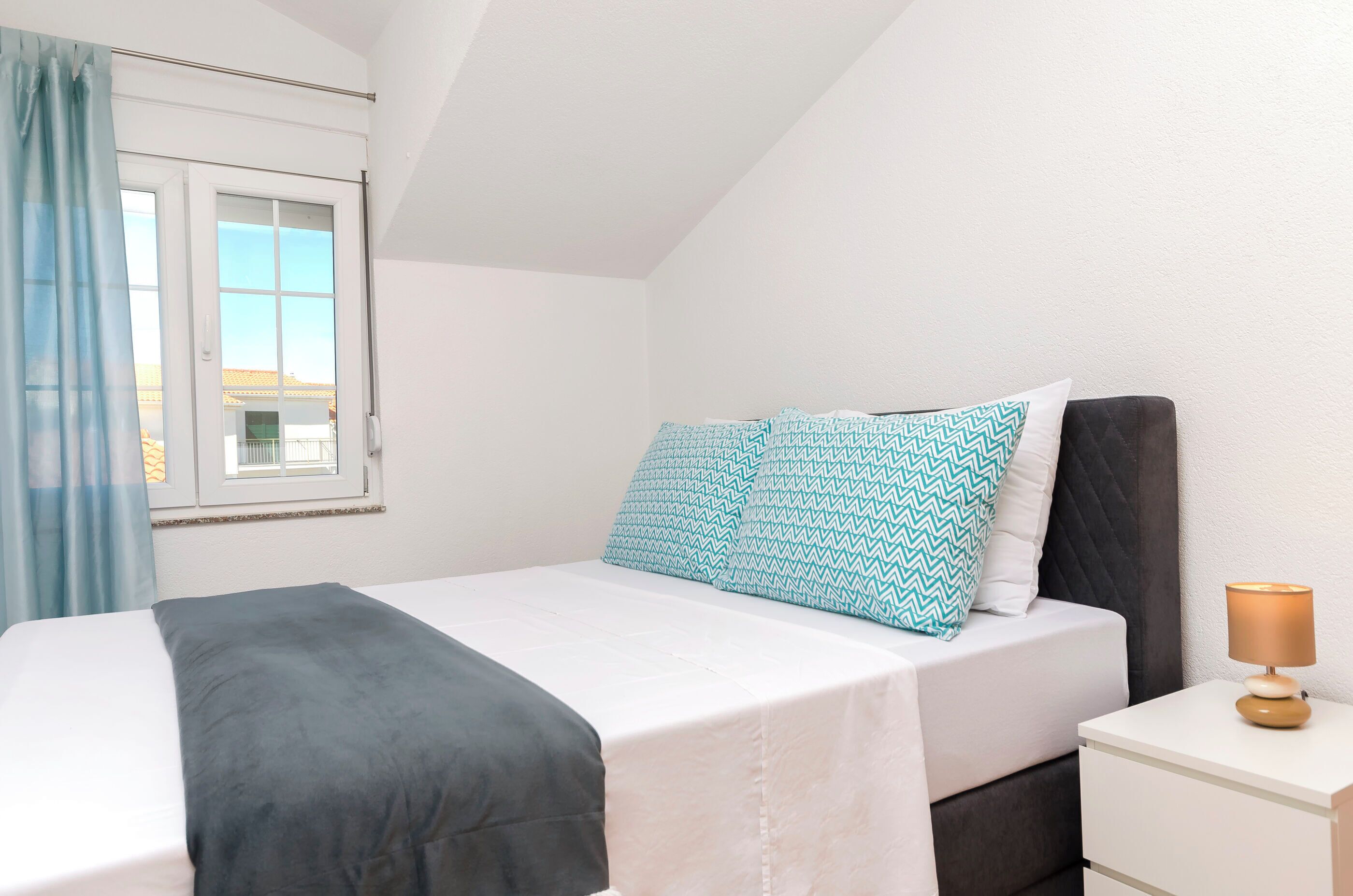 Quarto com cama, mesa de cabeceira, abajur e janela com cortinas.