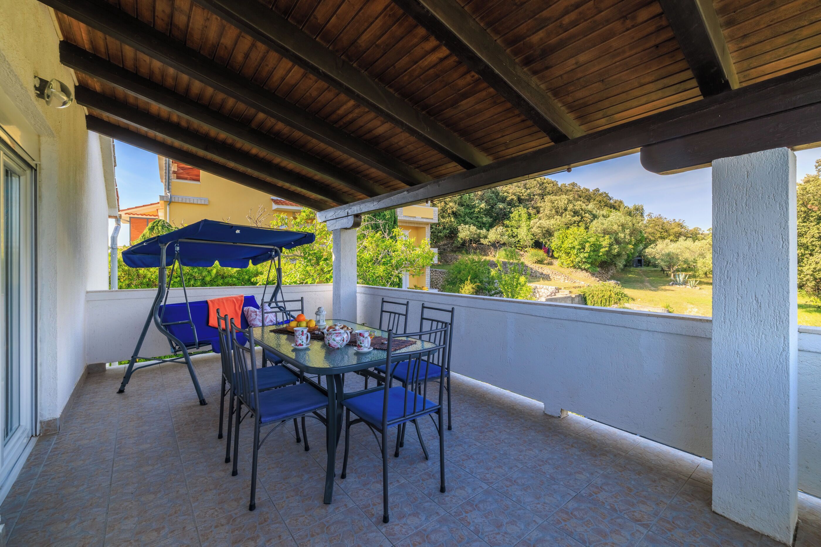 Um alpendre coberto com mesa e cadeiras, um guarda-sol azul e vista para uma encosta.