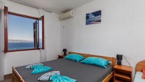 Um quarto com cama, duas mesas de cabeceira, um criado-mudo, janela com vista para o mar e um quadro na parede.
