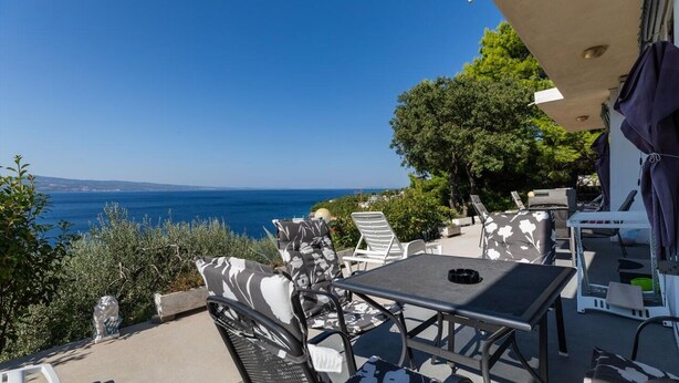 Um terraço ao ar livre com vista para o mar, com mesa e cadeiras, e uma área coberta também com mesa e cadeiras.