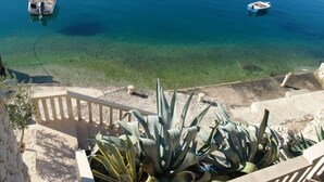 Vista costeira com um barco ancorado perto de um mar azul e límpido, uma escadaria descendo até à praia e uma grande planta de agave em primeiro plano.