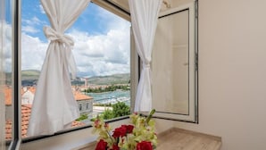 Quarto com vista para uma cidade costeira, janela com cortinas brancas e um vaso com flores vermelhas e amarelas sobre uma mesa de madeira.