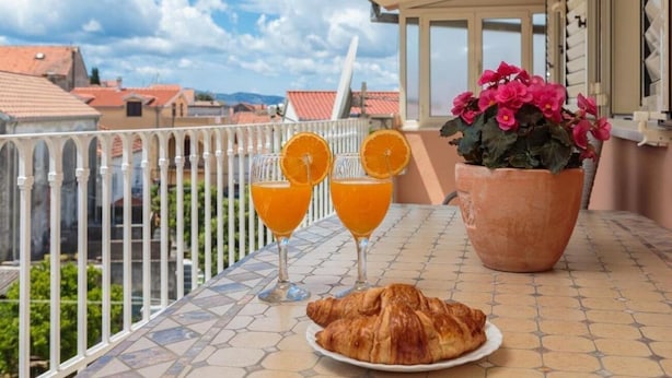 Uma varanda com chão de azulejos, guarda-corpo branco, uma mesa com um croissant, dois copos de sumo de laranja e um vaso com flores cor-de-rosa.