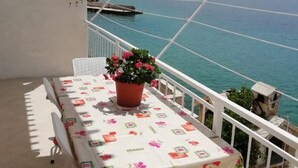 Uma varanda com mesa e cadeiras, um vaso de planta e vista para o mar e montanhas.