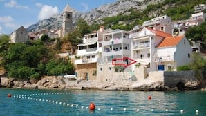 Uma vila costeira com edifícios ao longo da orla marítima, uma igreja e colinas rochosas ao fundo.