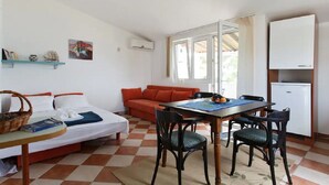 Quarto compacto com cama, sofá, mesa de jantar com cadeiras, pequena cozinha e janela que permite entrada de luz natural.