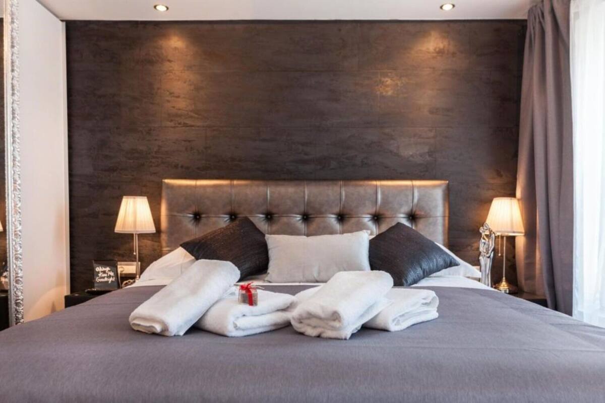 Uma cama bem arrumada com cabeceira estofada, duas luminárias de apoio e uma parede escura com textura.